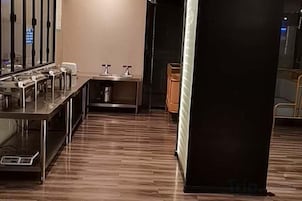 Quarto de hotel com piso de madeira, geladeira preta, uma mesa pequena com cadeira e janela com persianas.