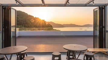 Terraço moderno ao ar livre com portas de vidro, oferecendo vista para um pôr do sol sobre a água e colinas.