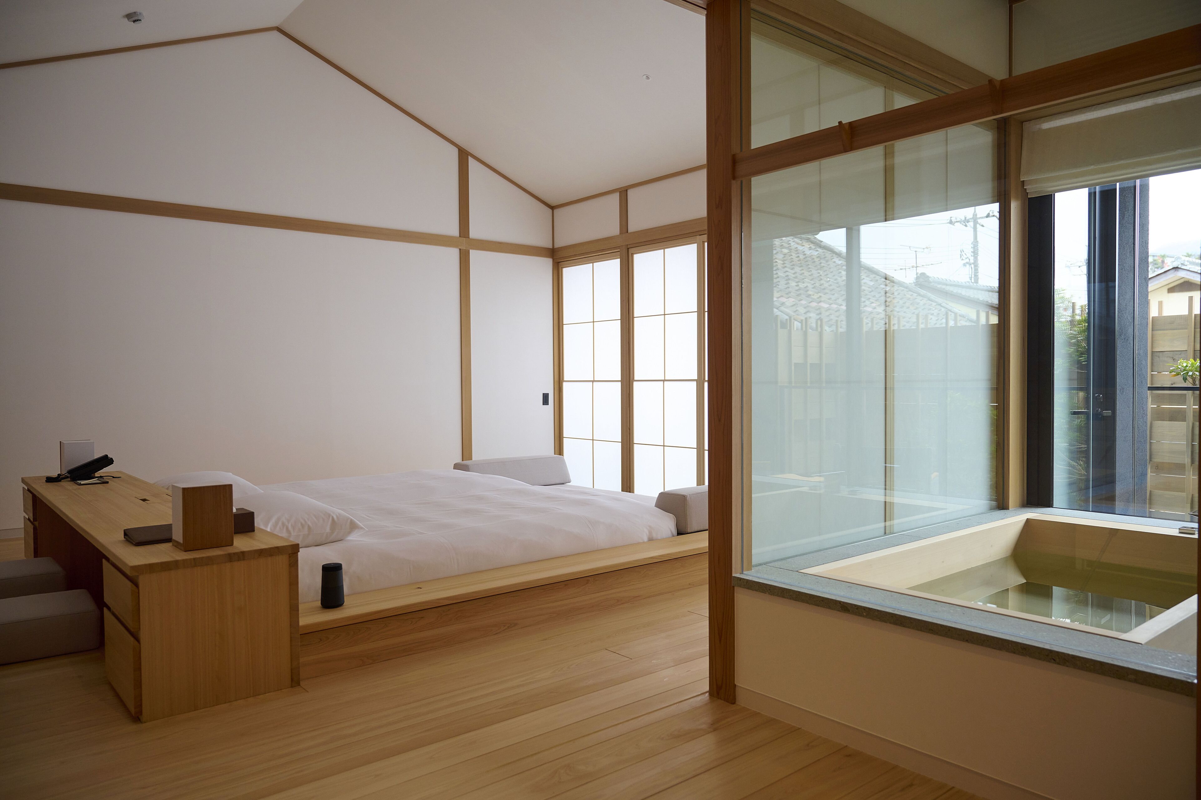 Quarto minimalista com escrivaninha de madeira, cama e uma janela grande.