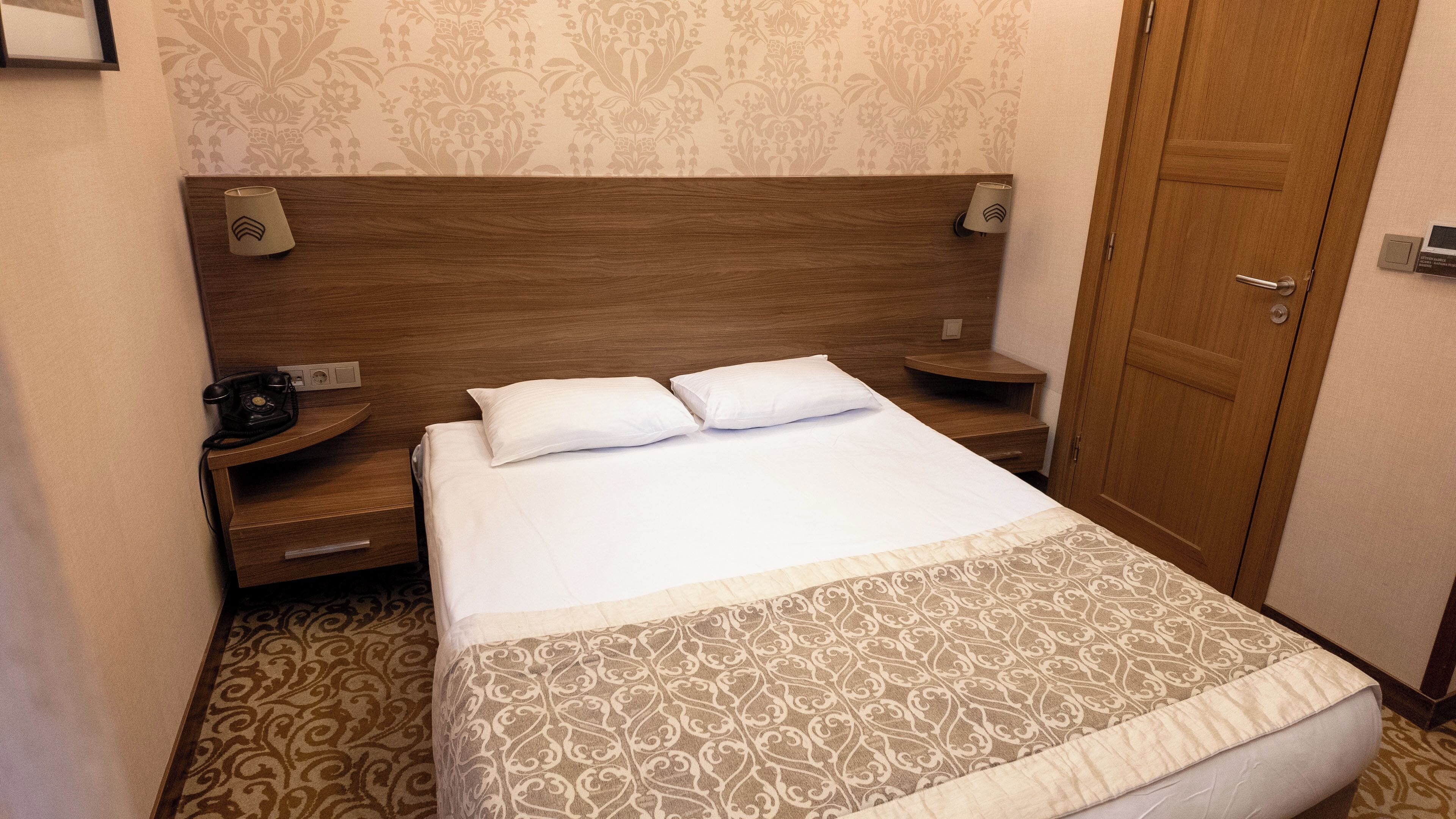 Quarto de hotel com cabeceira de madeira, cama com lençóis brancos, duas mesas de cabeceira com luminárias, um telefone e um guarda-roupa de madeira.