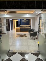 Uma área de recepção de hotel, com balcão, sala de espera com cadeiras e piso estampado.