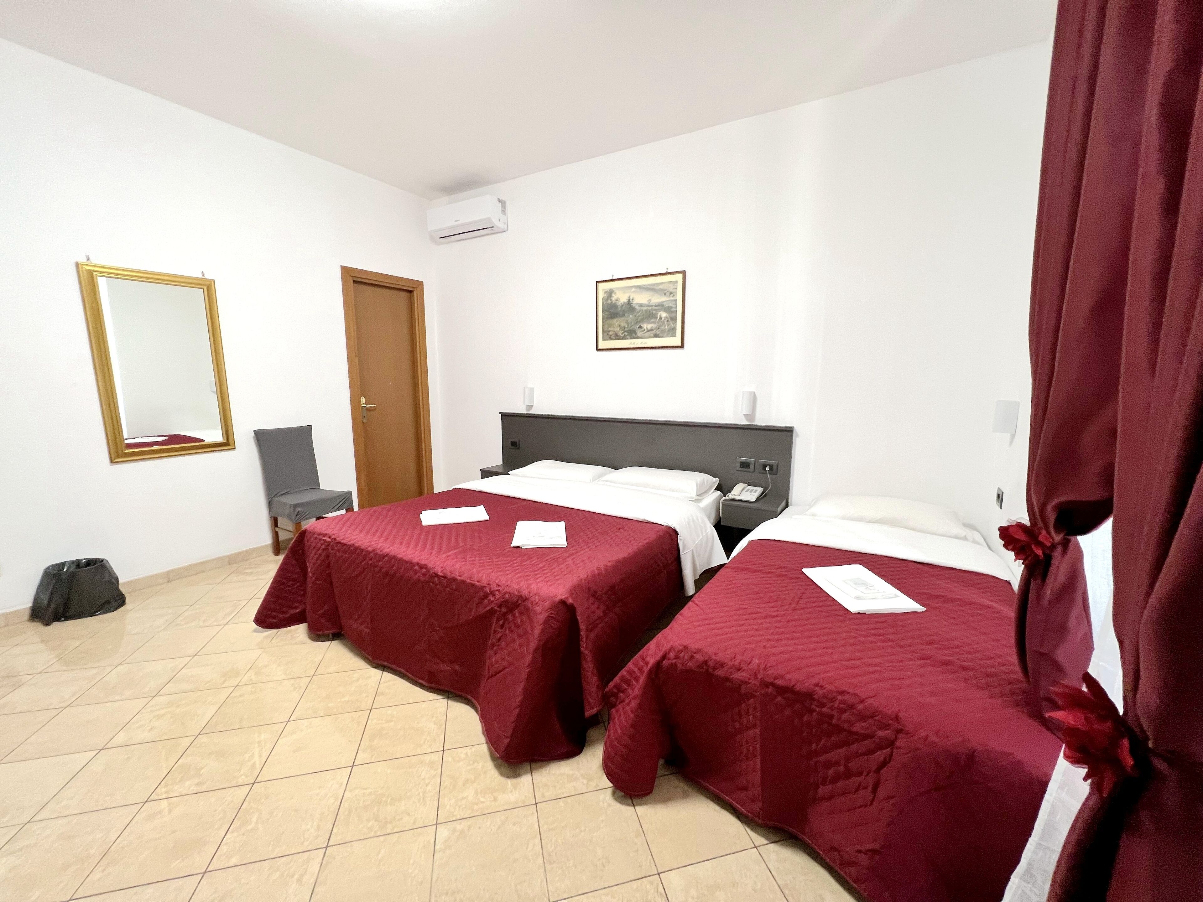 Quarto de hotel com duas camas, uma escrivaninha, uma cadeira, um espelho e uma porta.