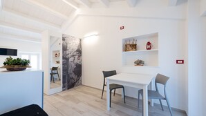 Quarto moderno e bem iluminado, com uma mesa e cadeiras brancas, uma mala preta e um vaso de planta.