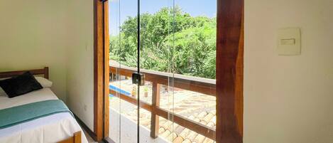 Quarto com cama, varanda com guarda-corpo de madeira e vista para o verde lá fora.
