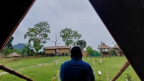 Uma pessoa dentro de uma estrutura tipo tenda, com vista para uma área gramada e edifícios ao fundo.