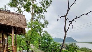 Uma casa rústica de madeira com telhado de palha, cerca de bambu e um caminho de cascalho que leva a um corpo d'água, com montanhas ao fundo.