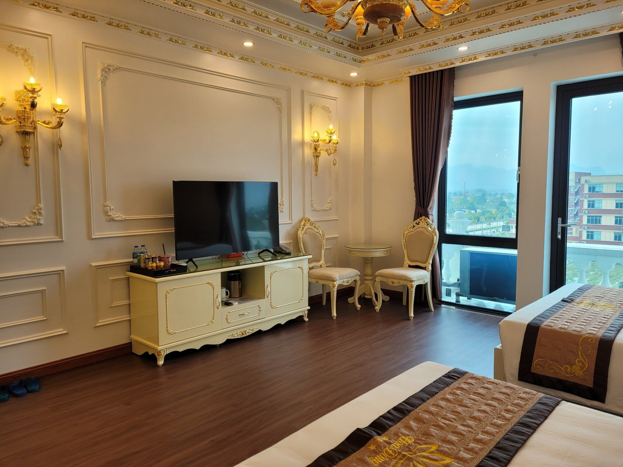 Quarto de hotel com uma televisão de tela plana grande, um armário branco ornamentado, duas cadeiras brancas, uma mesa redonda pequena e uma cama grande com colcha estampada.