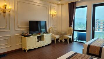 Quarto de hotel com uma televisão de tela plana grande, um armário branco ornamentado, duas cadeiras brancas, uma mesa redonda pequena e uma cama grande com colcha estampada.