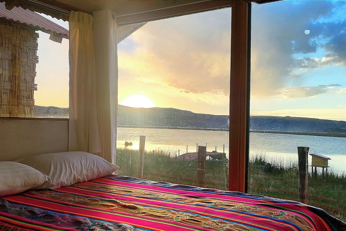 Quarto com uma janela grande que oferece vista para um lago e montanhas ao longe.