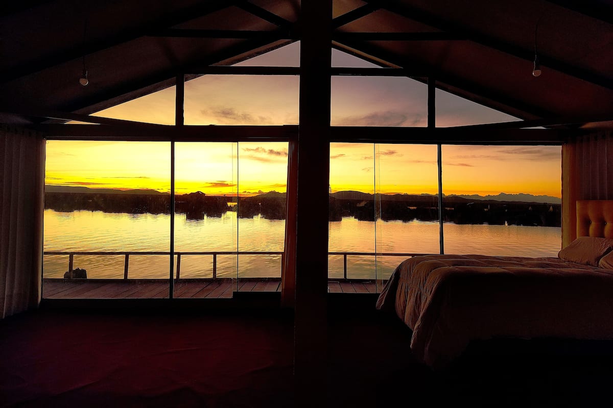 Quarto com uma grande janela com vista para um corpo d'água ao pôr do sol.
