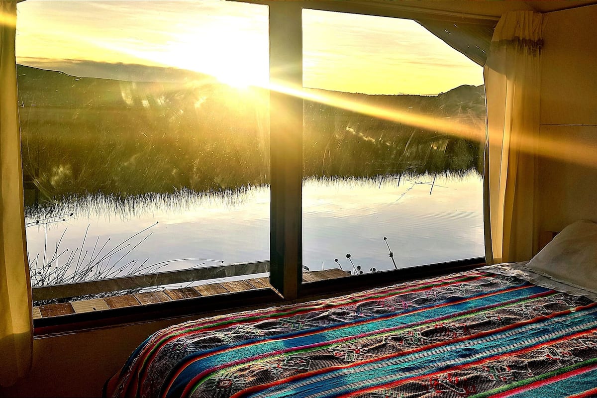 Quarto com cama e uma grande janela com vista para um lago ao pôr do sol.
