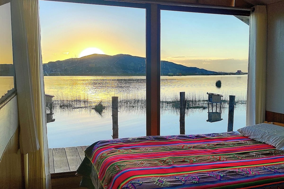 Quarto com uma grande janela com vista para um lago e montanhas ao pôr do sol.