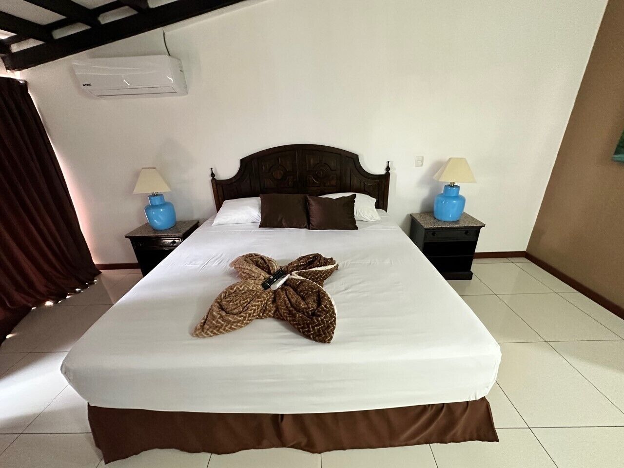 Cama bem arrumada com manta marrom, duas mesas de cabeceira com abajures e um ar condicionado na parede.
