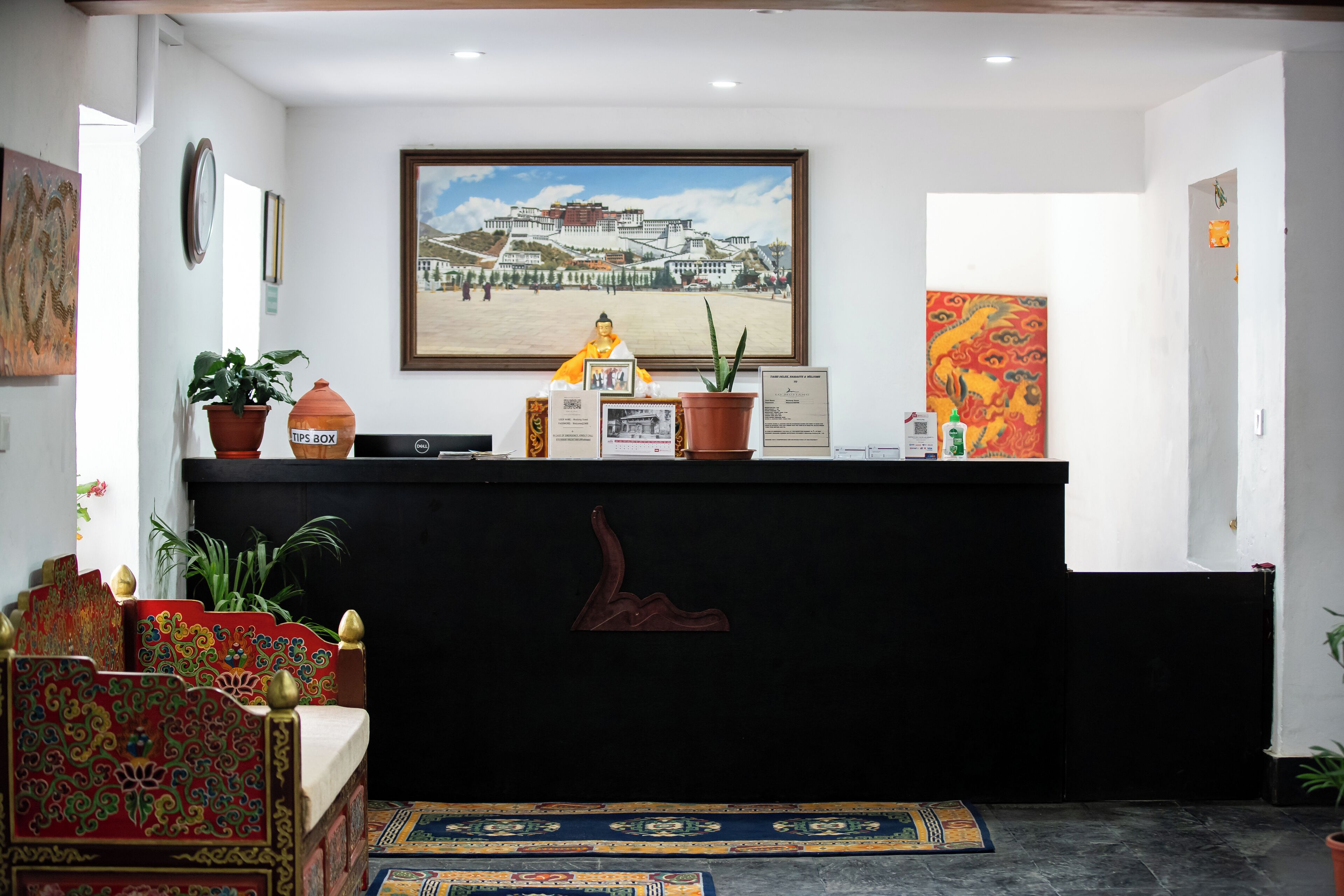 Uma área de recepção com balcão preto, quadro de um edifício, plantas em vasos e decoração tradicional.