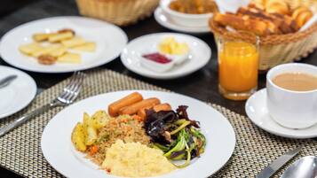 Uma mesa de café da manhã com pratos de comida, uma cesta de pão, um copo de suco de laranja e uma xícara de café.