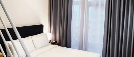 Quarto de hotel moderno com uma cama bem arrumada, uma escada com degraus de madeira e uma grande janela com cortinas.