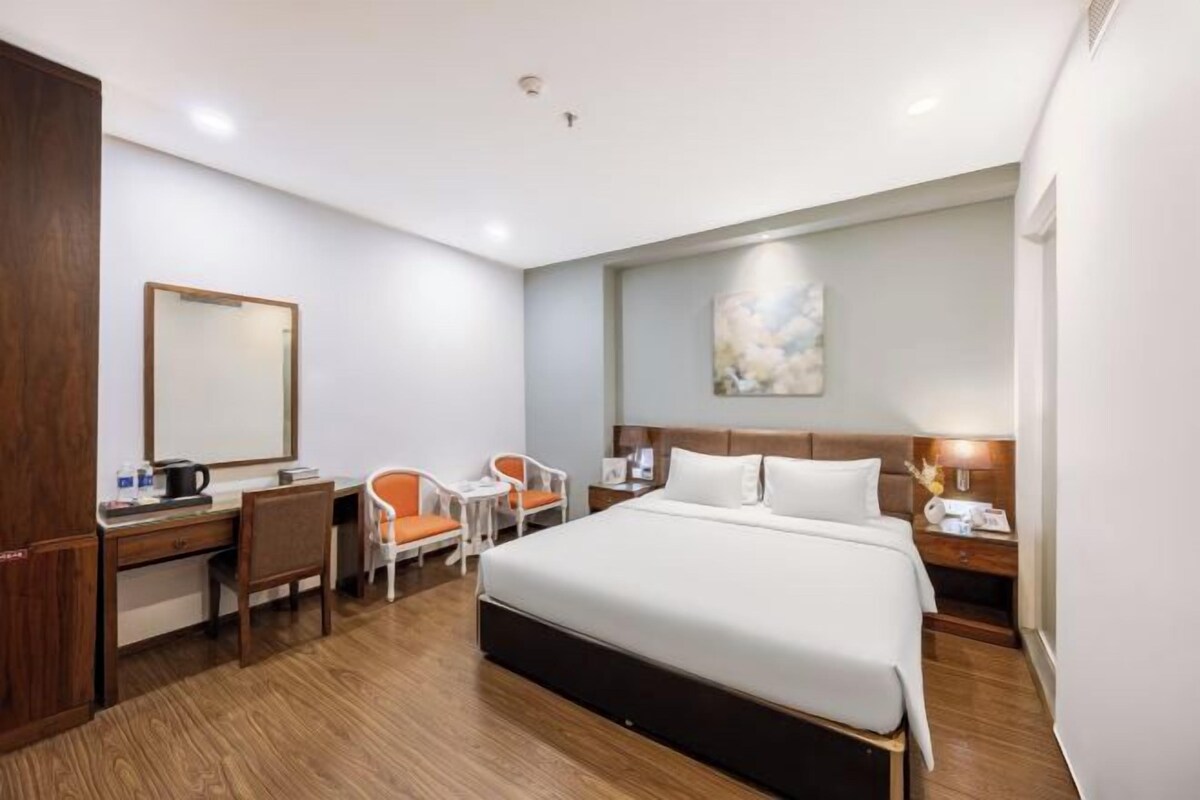 Quarto de hotel com uma cama grande, uma escrivaninha com cadeira, uma mesinha e uma mesa de cabeceira.
