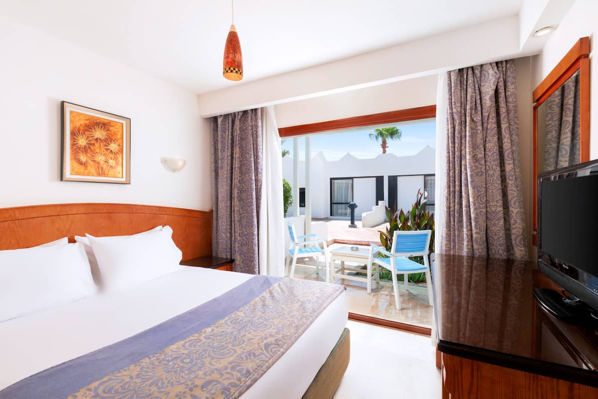 Quarto de hotel com uma cama grande, televisão e varanda com cadeiras e mesa.