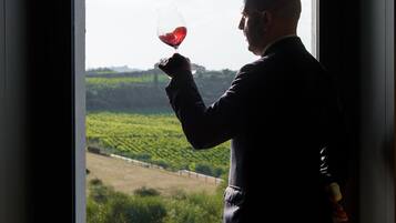Homem de terno segurando uma taça de vinho, olhando para um vinhedo pela janela.