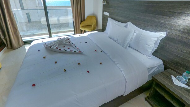 Quarto de hotel com uma cama grande, cabeceira de madeira, poltrona amarela e vista para o mar através de grandes janelas.