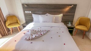 Quarto de hotel com uma cama grande, duas poltronas amarelas, uma cabeceira de madeira e uma mesinha de cabeceira com uma garrafa de água.