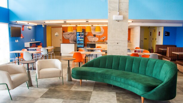 Uma área de lounge moderna com um sofá verde, cadeiras laranja e mesas. Há máquinas de venda automática e uma tela grande ao fundo.