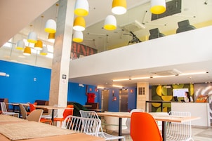 Restaurante moderno com pé-direito alto, luminárias pendentes amarelas e brancas, e uma combinação de cadeiras laranja e brancas.