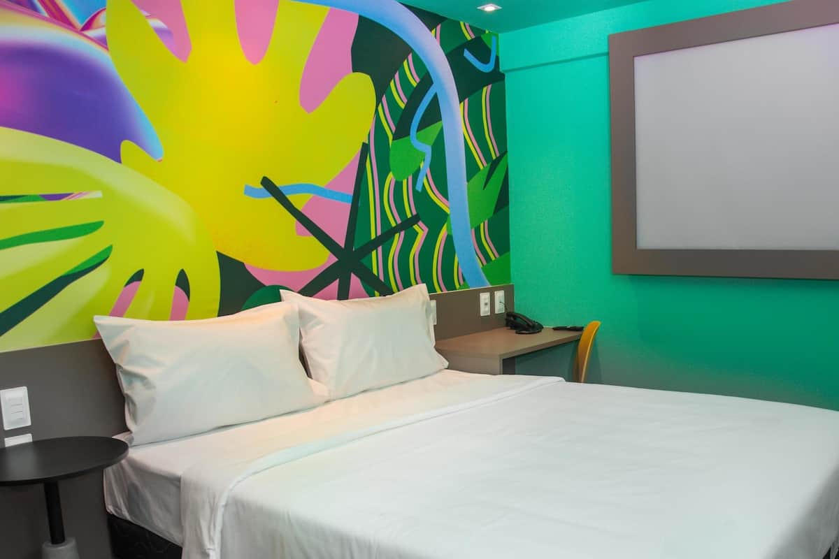 Quarto de hotel com cama, escrivaninha, cadeira e um mural de parede colorido.