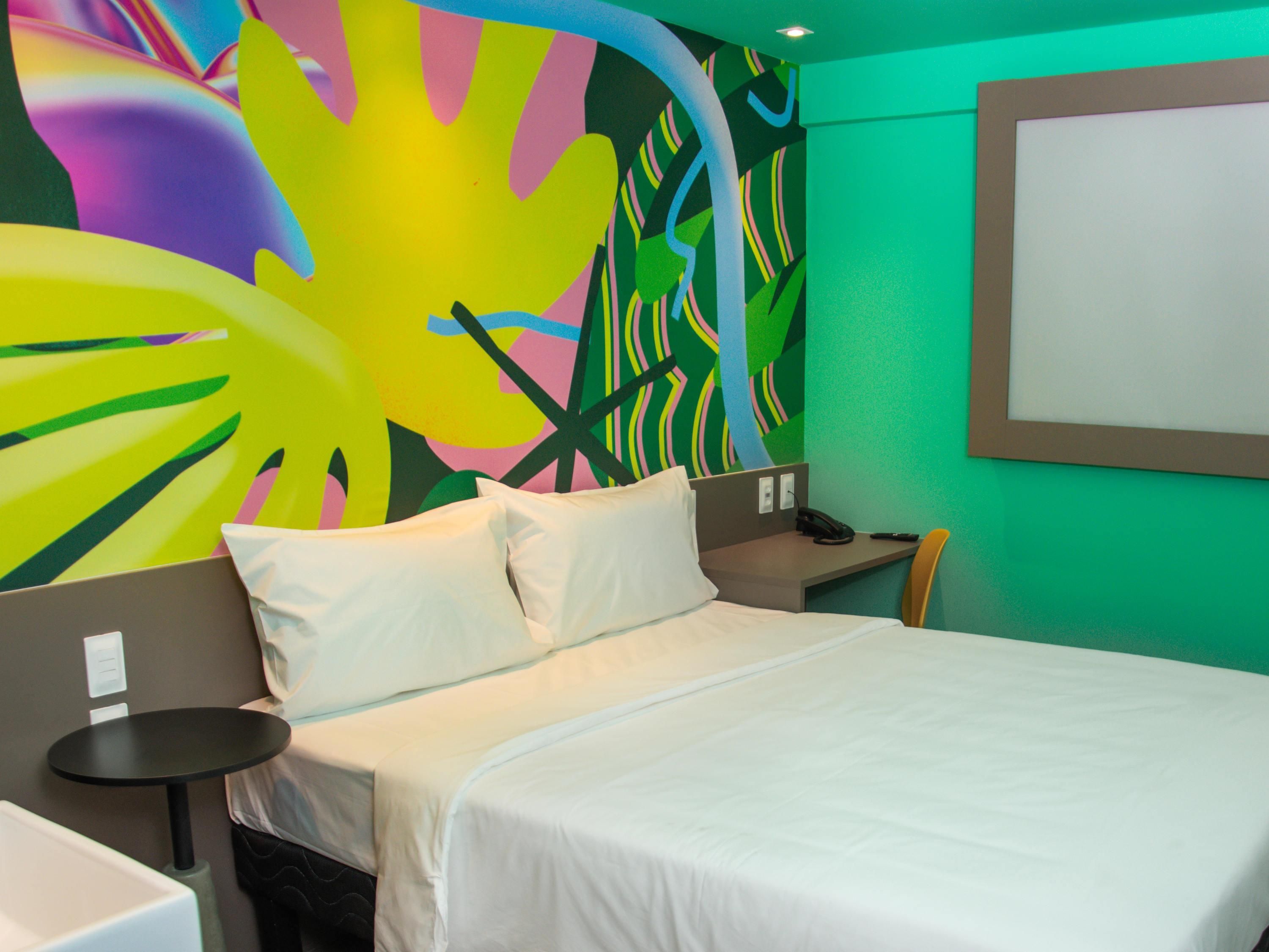 Quarto de hotel com cama, escrivaninha, cadeira, telefone e um grande mural abstrato na parede.