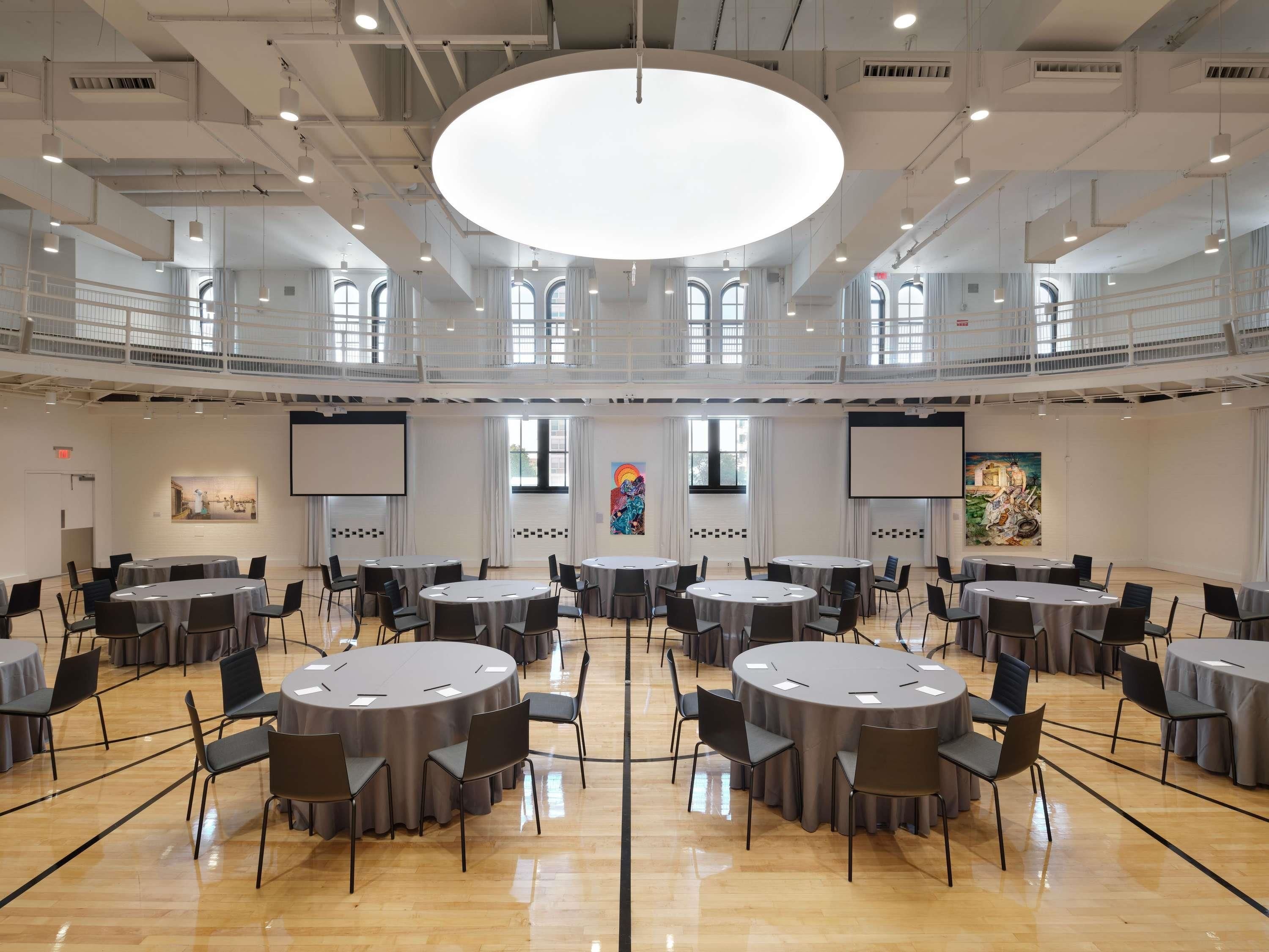 Uma sala de conferências ampla, com várias mesas e cadeiras, janelas grandes e um teto alto com vigas aparentes.