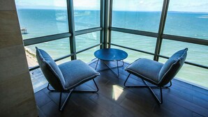 Quarto moderno com amplas janelas com vista para um corpo de água. Há duas poltronas cinzas e uma pequena mesa redonda.