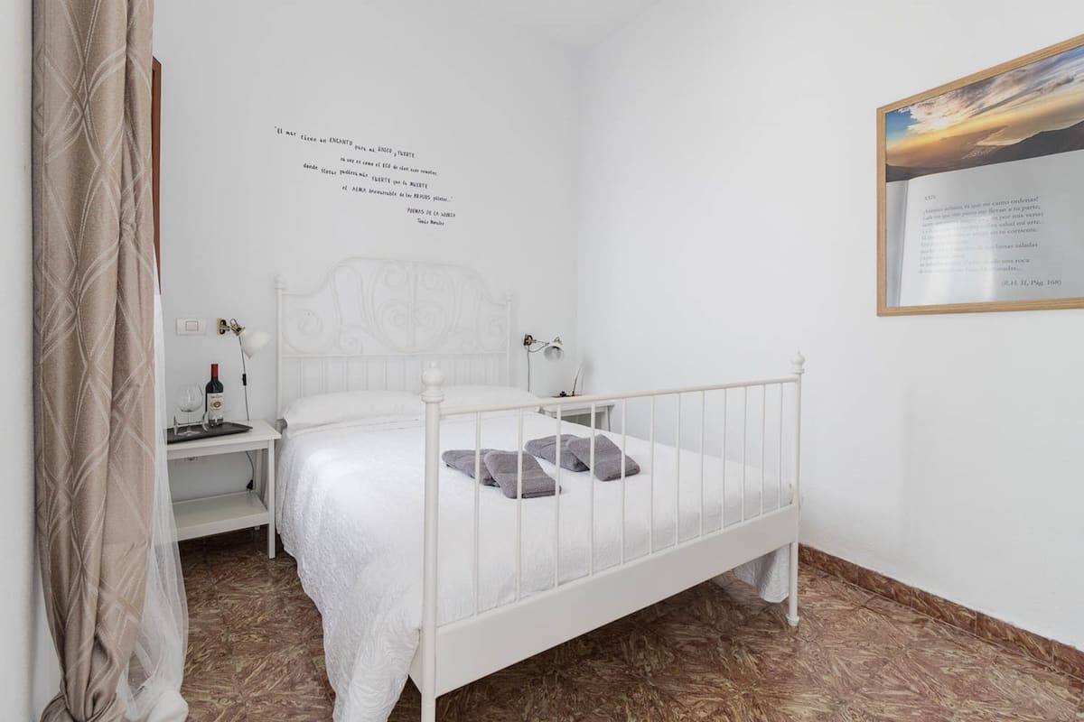 Quarto branco com cama, mesa de cabeceira, abajur e parede com texto e uma moldura.