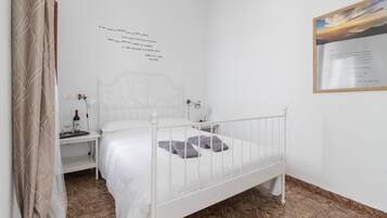 Quarto branco com cama, mesa de cabeceira, abajur e parede com texto e uma moldura.
