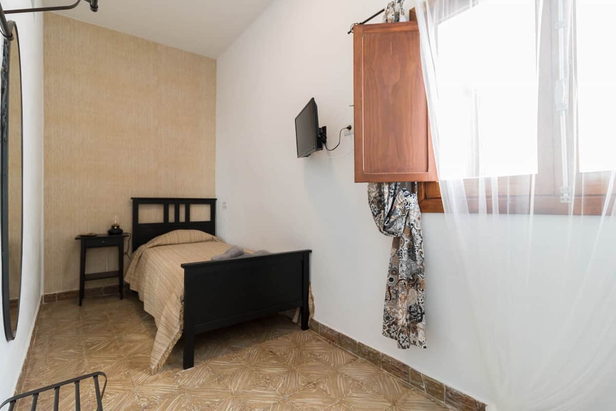 Quarto com cama, mesa de cabeceira, TV fixada na parede e janela com cortinas leves.