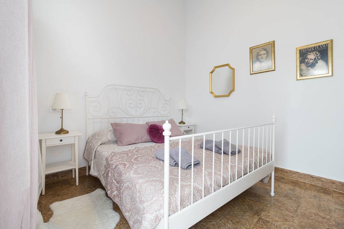 Quarto com estrutura de cama de metal branco, colcha estampada, duas mesas de cabeceira com abajures e um tapete no chão.