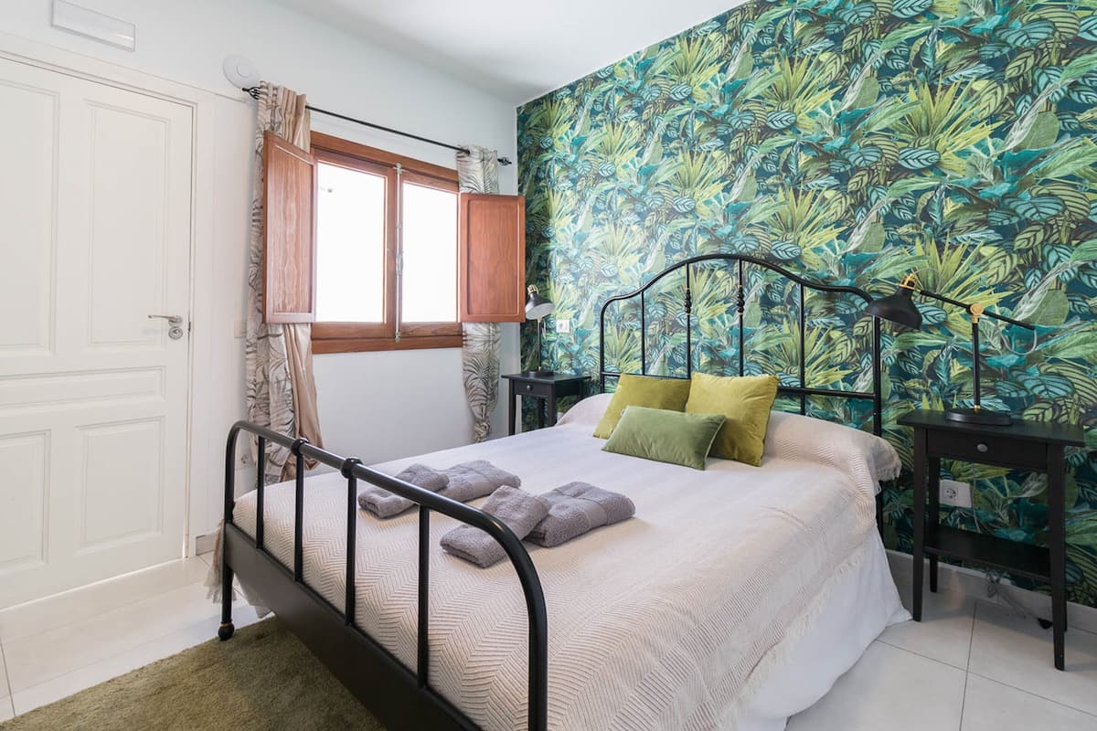 Quarto com cama, cortinas, janela, mesa de cabeceira e parede com padrão de folhas.