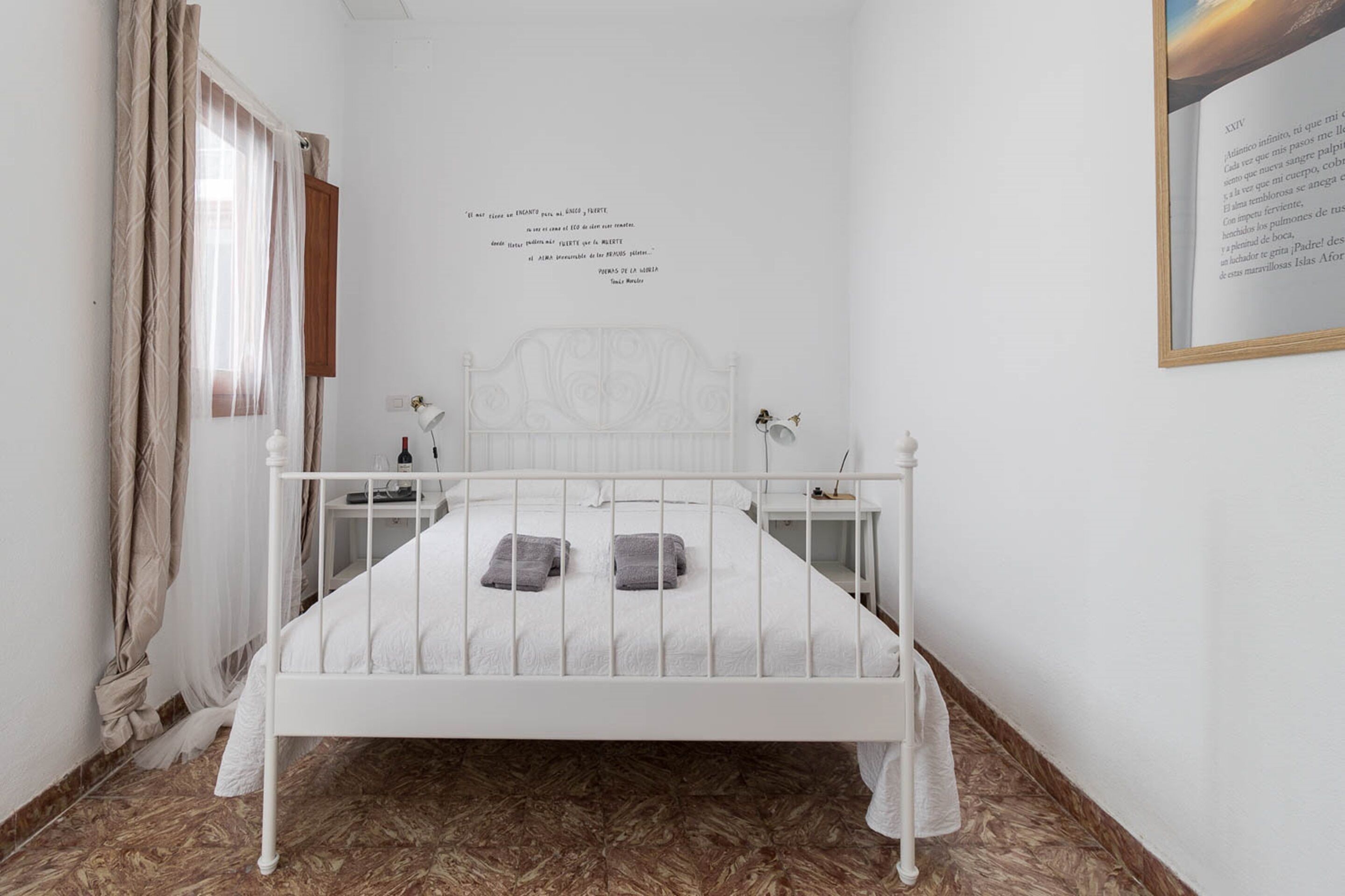 Uma cama de metal branca com duas almofadas, uma mesa de cabeceira com abajur e uma parede com um texto emoldurado.