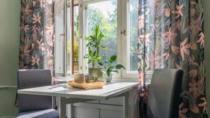 Uma área de refeições com uma mesa, duas cadeiras, um vaso de planta e uma janela com cortinas florais.