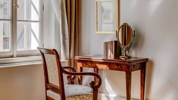 Um quarto com piso de parquet de madeira, uma cadeira com assento acolchoado, uma mesa pequena com um espelho e uma janela com cortinas leves.