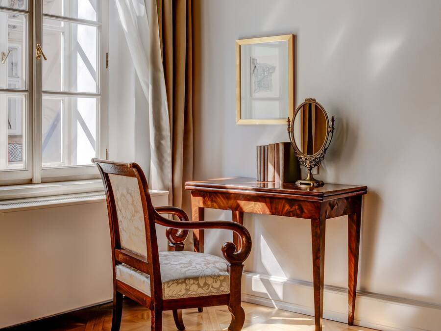 Um quarto com piso de parquet de madeira, uma cadeira com assento acolchoado, uma mesa pequena com um espelho e uma janela com cortinas leves.