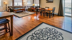 Uma sala de estar ampla com piso de madeira, um conjunto de sofás, uma mesa de jantar com cadeiras e uma grande janela com vista.