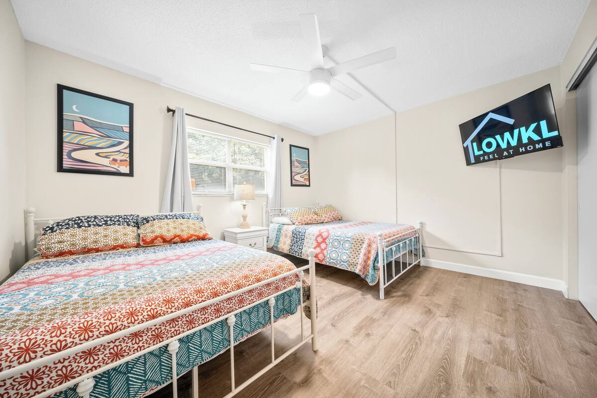 Quarto com duas camas, ventilador de teto e uma TV exibindo 'LOWKL FEEL AT HOME'.