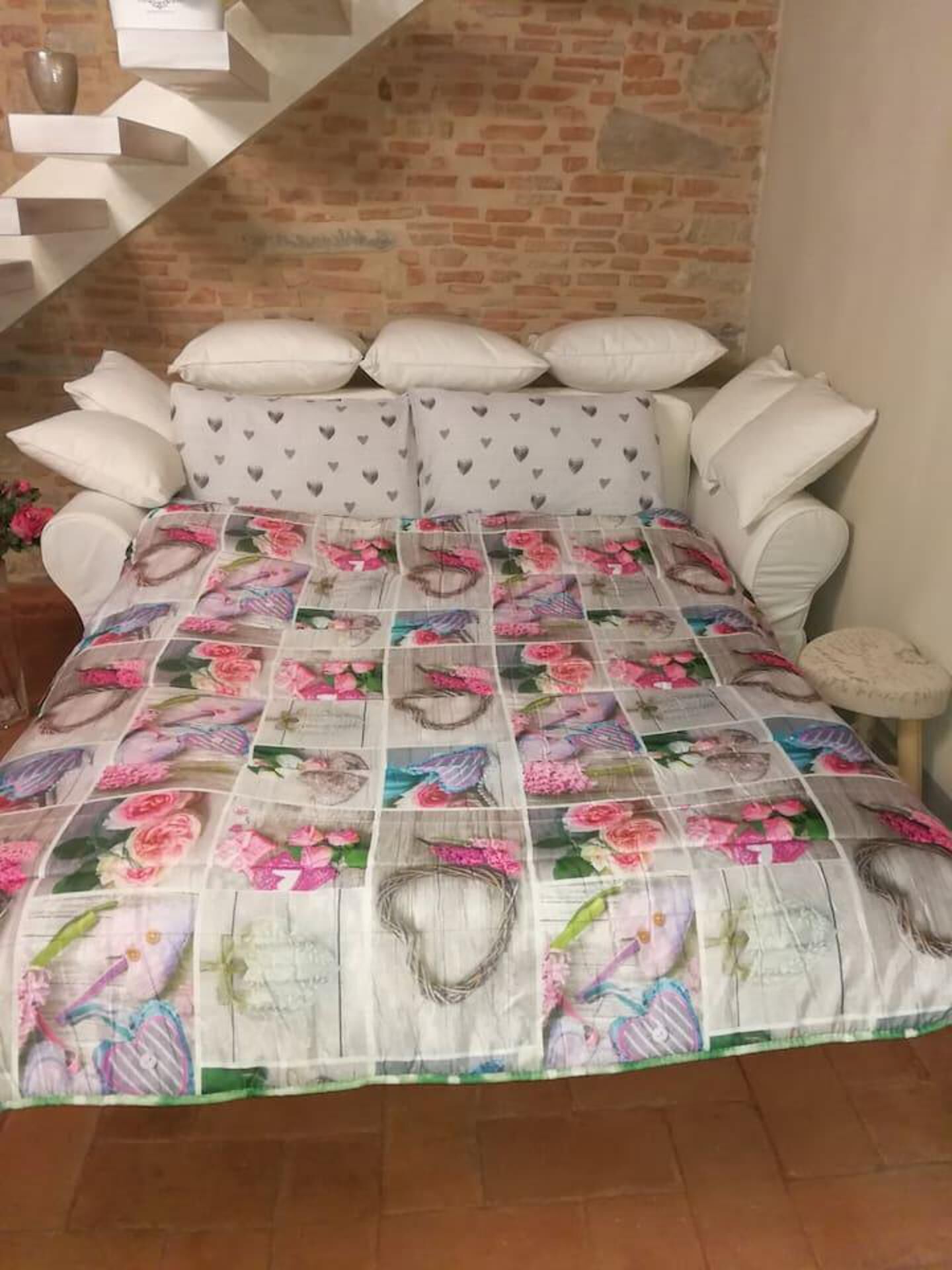 Cama com colcha floral, travesseiros brancos e parede de tijolos ao fundo.