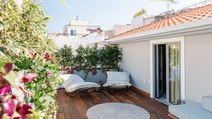 Um terraço no telhado com piso de madeira, duas cadeiras brancas com almofadas, um tapete redondo tecido e uma pequena mesa com uma planta em vaso.
