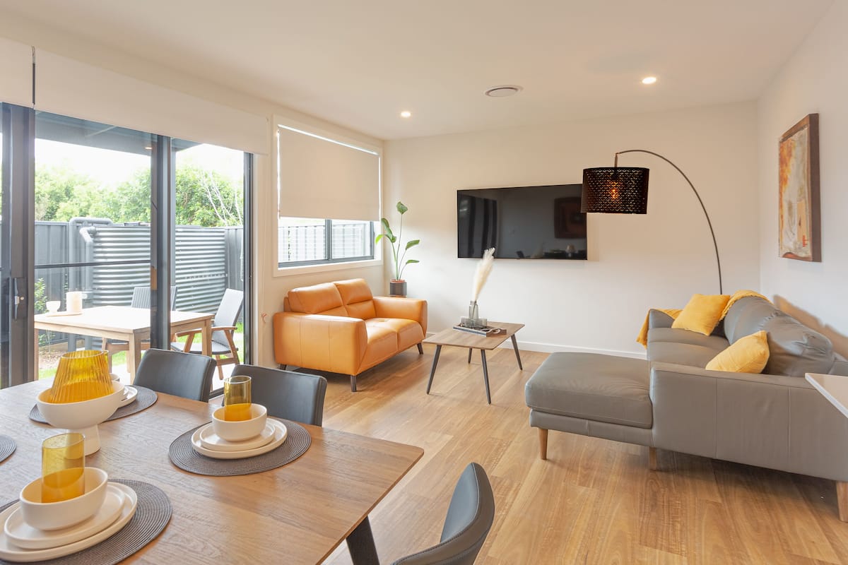 Uma sala de estar moderna com área de jantar, um sofá cinza e uma TV fixada na parede.