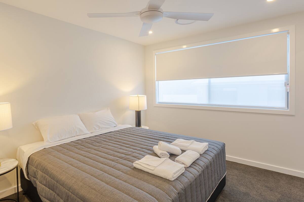 Quarto de hotel com cama, duas luminárias de cabeceira, ventoinha no teto, janela com persianas e cama bem arrumada com toalhas.