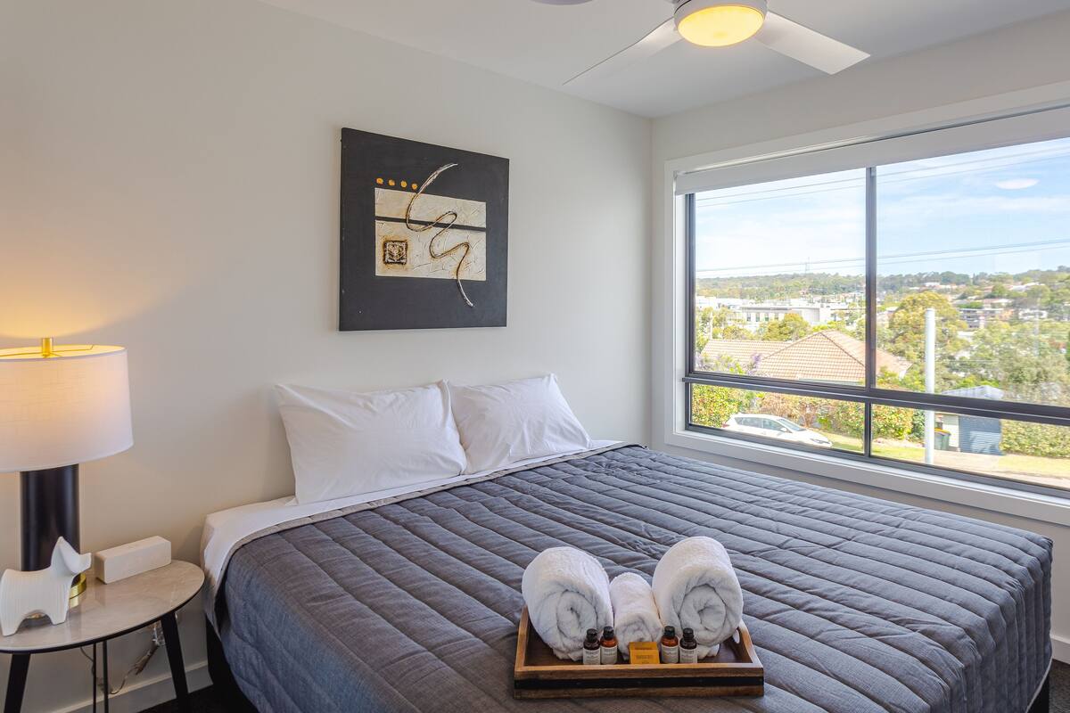 Um quarto com uma cama grande, uma mesinha de cabeceira, um abajur, um quadro e uma janela com vista para a cidade.