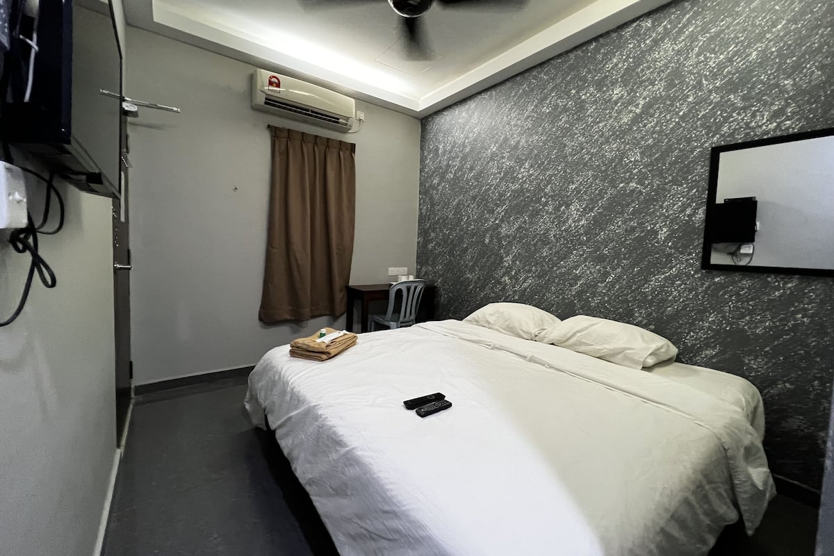 Quarto de hotel com cama, escrivaninha, ar condicionado, ventoinha no teto e televisão fixada na parede.