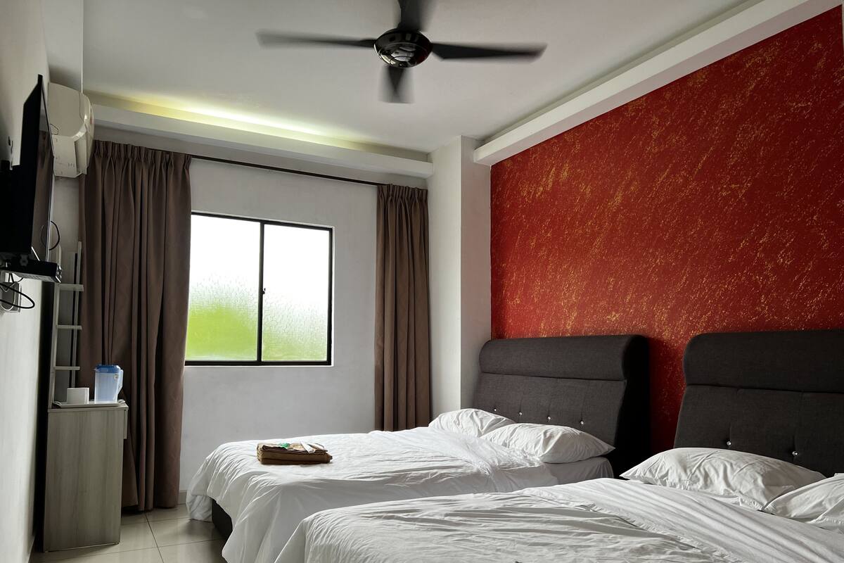 Quarto de hotel com duas camas, ventoinha no teto, janela com cortinas, parede vermelha com textura e uma pequena mesa com uma garrafa de água.