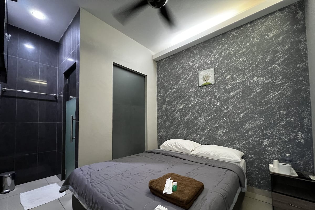 Quarto moderno com cama cinza, uma pequena mesa de cabeceira, um criado-mudo, parede texturizada cinza, uma porta e área de chuveiro.